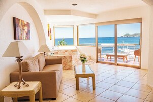 Uma sala de estar ampla, com um sofá, uma mesa de centro e vista para o mar através de grandes janelas.