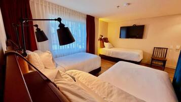 Quarto de hotel com duas camas, uma mesa, uma cadeira, televisão e janela com cortinas.
