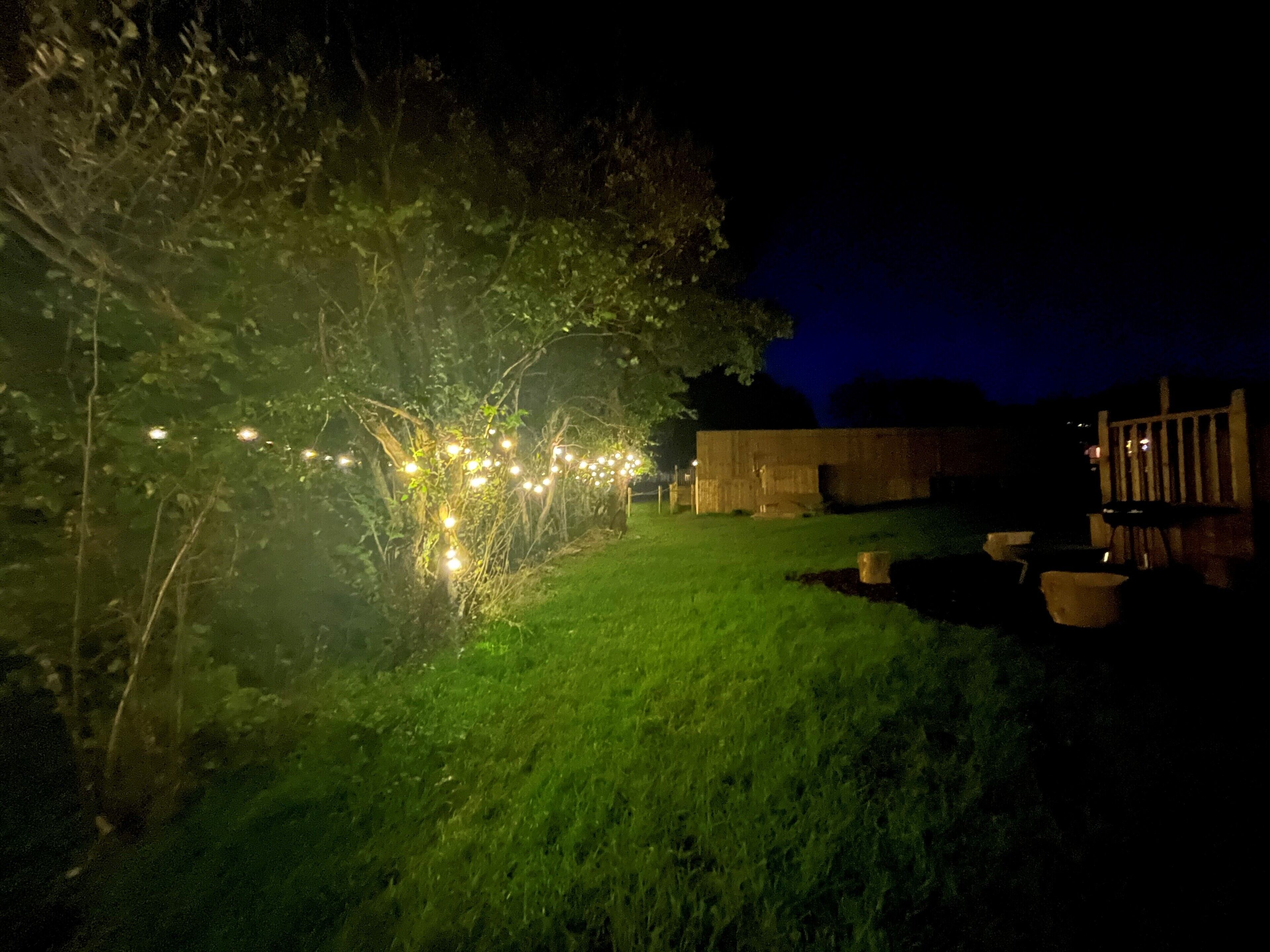 Área externa iluminada com luzes de corda, gramado e uma estrutura de madeira.