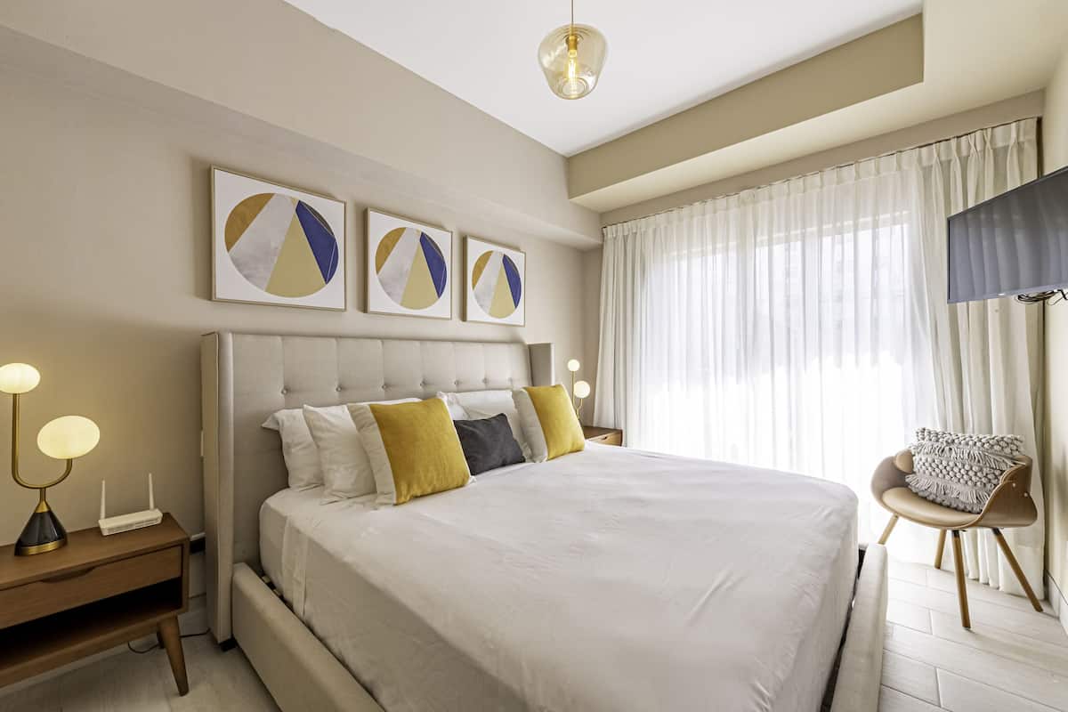 Um quarto moderno com uma cama grande, uma mesa de cabeceira com luminária, uma cadeira e obras de arte na parede.