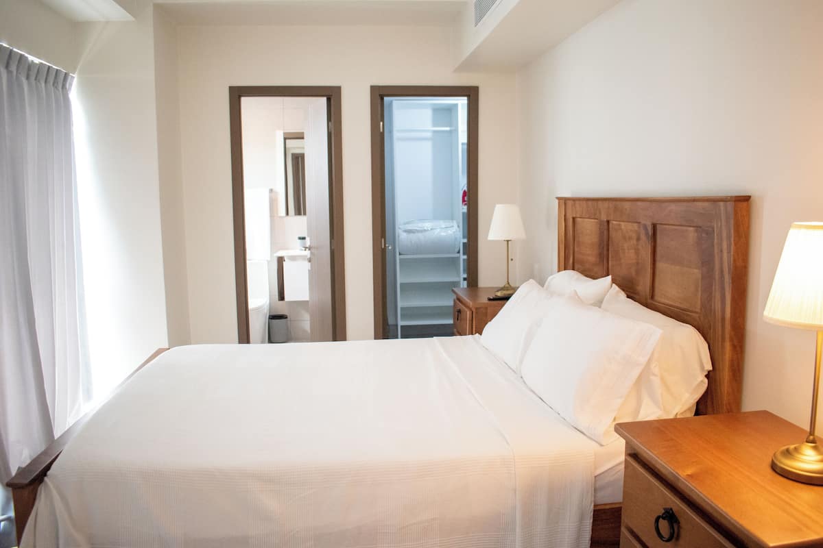 Quarto de hotel com uma cama grande, uma mesinha de cabeceira de madeira com abajur, um guarda-roupa e um banheiro visível por uma porta aberta.