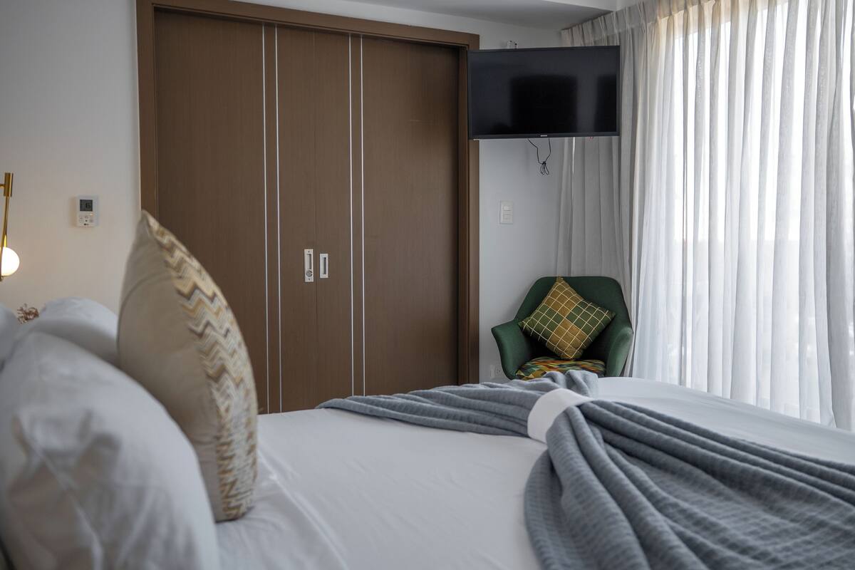 Quarto de hotel com uma cama, uma poltrona verde, uma televisão e um guarda-roupa.