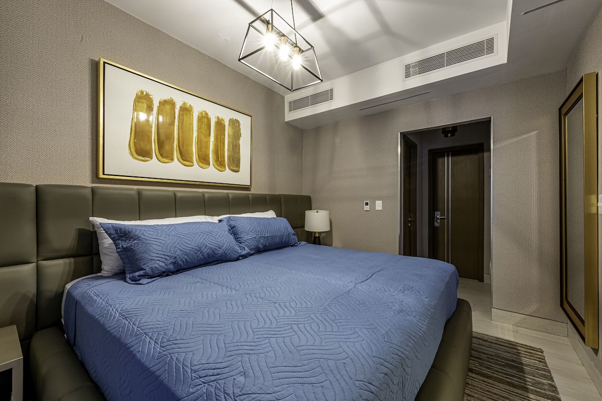 Quarto de hotel moderno com uma cama grande, uma obra de arte emoldurada acima da cama, uma mesa de cabeceira, um abajur e uma porta.