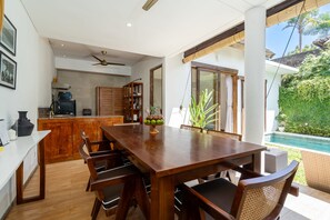 Uma sala de jantar com mesa e cadeiras de madeira, cozinha ao fundo e vista para uma piscina.