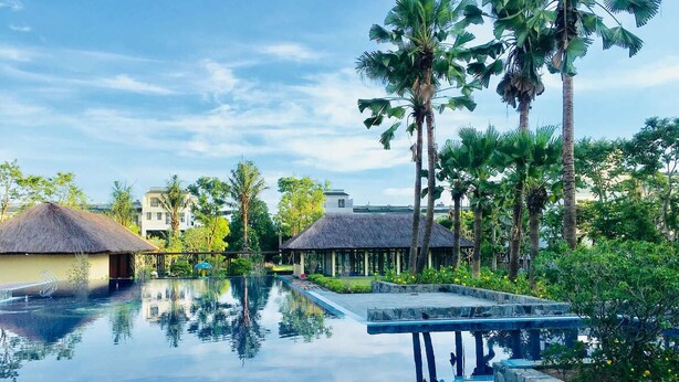 Piscina cercada por palmeiras e estruturas com telhados de palha, provavelmente parte de um resort.