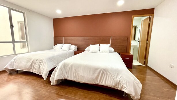 Quarto de hotel com duas camas, cabeceiras de madeira, chão de madeira, uma janela e uma porta que dá para um banheiro.