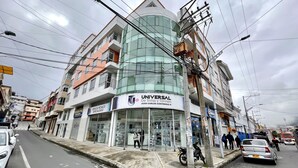 Um edifício de vários andares com fachada de vidro, uma vitrine visível com placa da 'Universal De Tintas y Toners', e uma motocicleta estacionada na calçada.