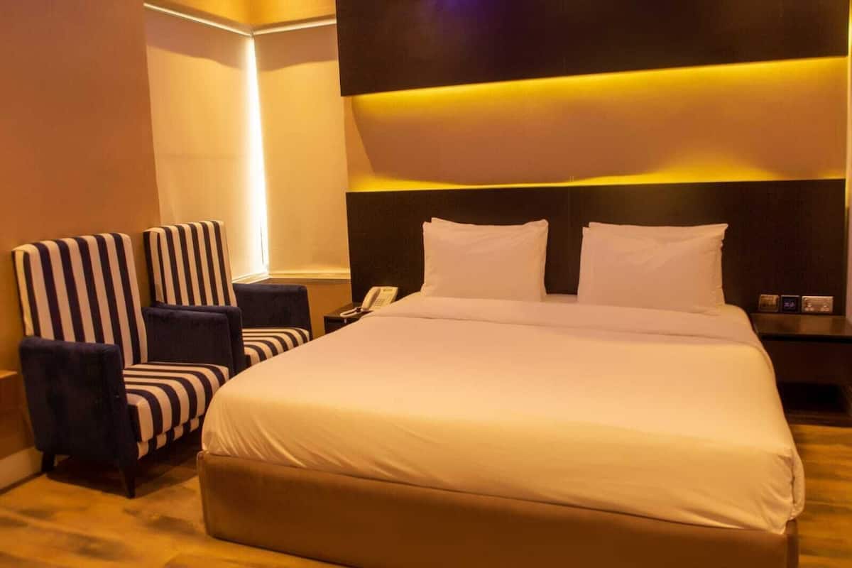 Quarto de hotel com uma cama grande, duas poltronas listradas, uma mesa de cabeceira com telefone e um criado-mudo com relógio.