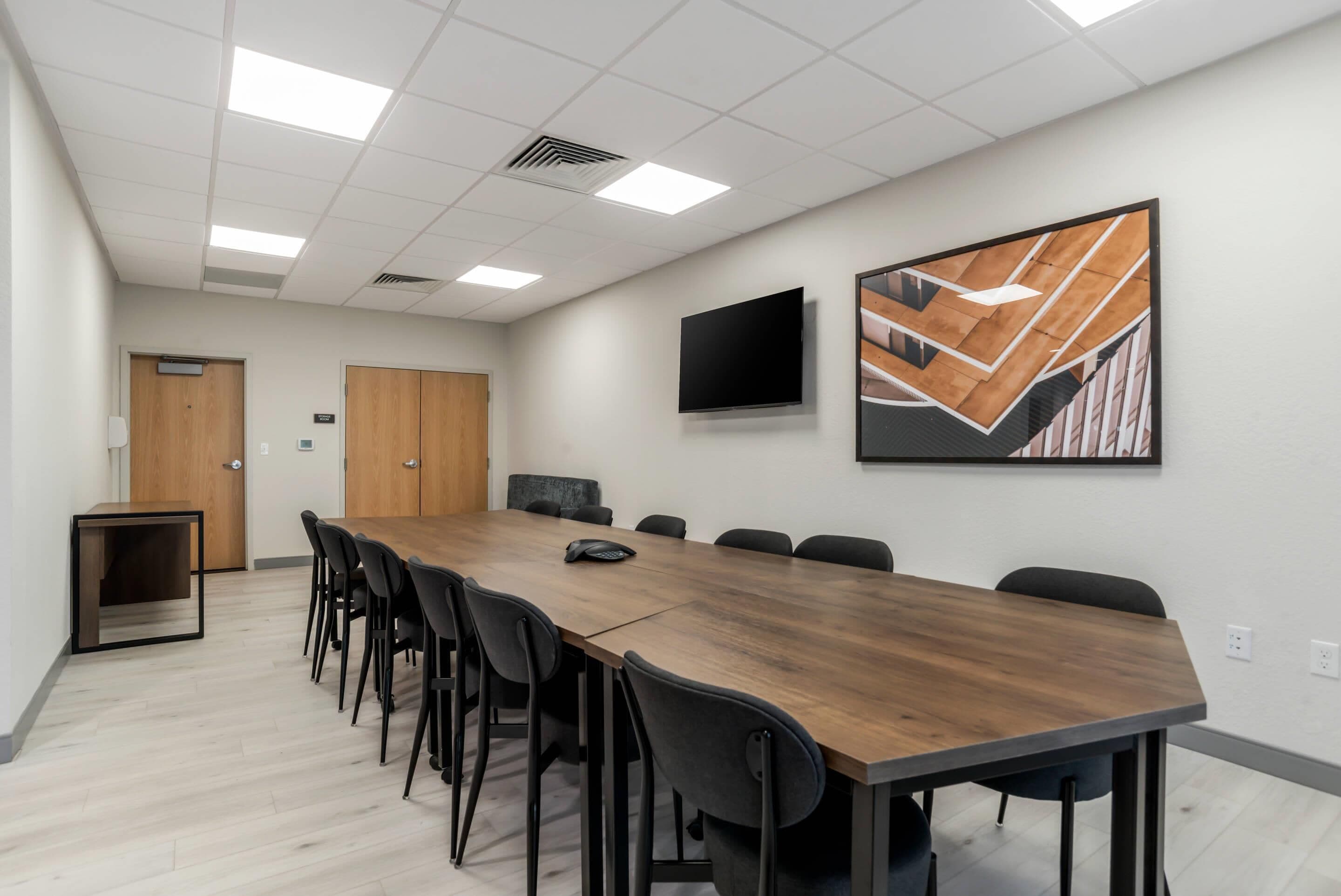 Sala de conferências com uma mesa comprida, cadeiras, uma TV de tela plana e uma obra de arte abstrata emoldurada na parede.