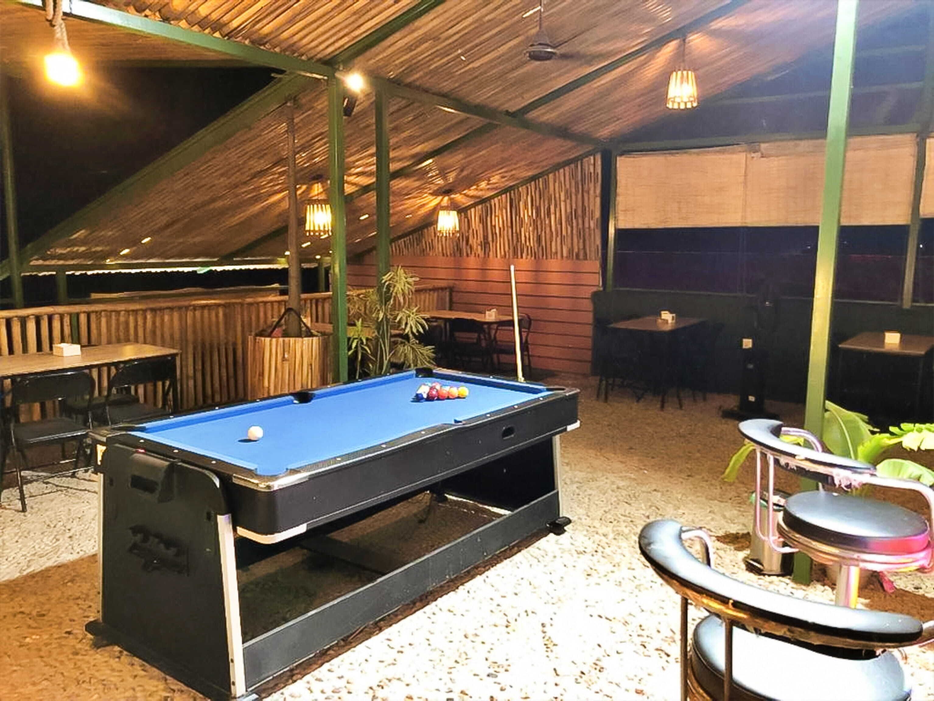 Uma mesa de bilhar coberta por um feltro azul, rodeada por mesas e cadeiras, sob um teto de madeira com luzes pendentes.