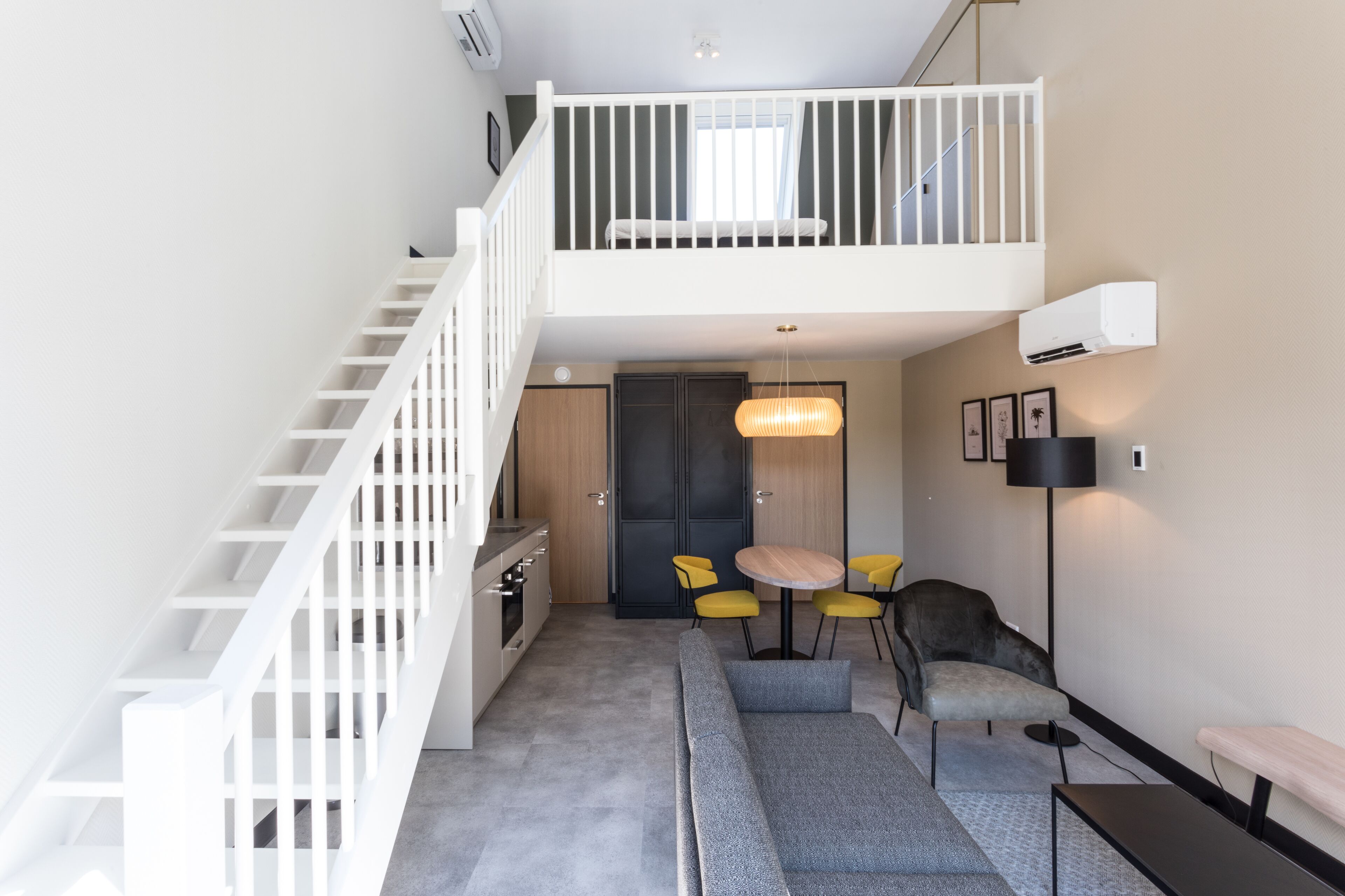 Um apartamento moderno com uma escada que dá acesso a um dormitório no mezanino, uma área de jantar com cadeiras amarelas e um sofá cinza.
