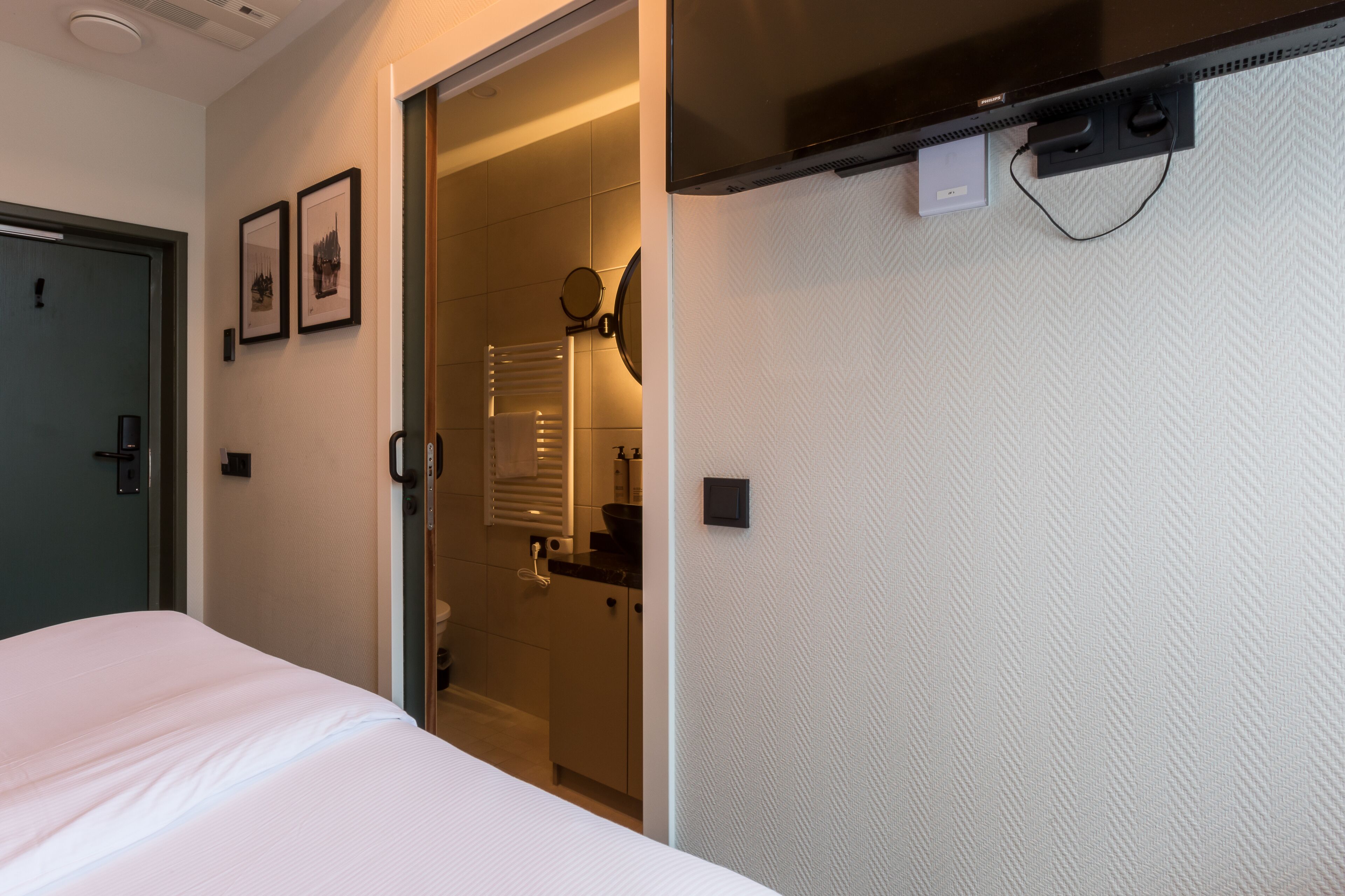 Quarto de hotel com cama, porta, espelho e televisão presa à parede.