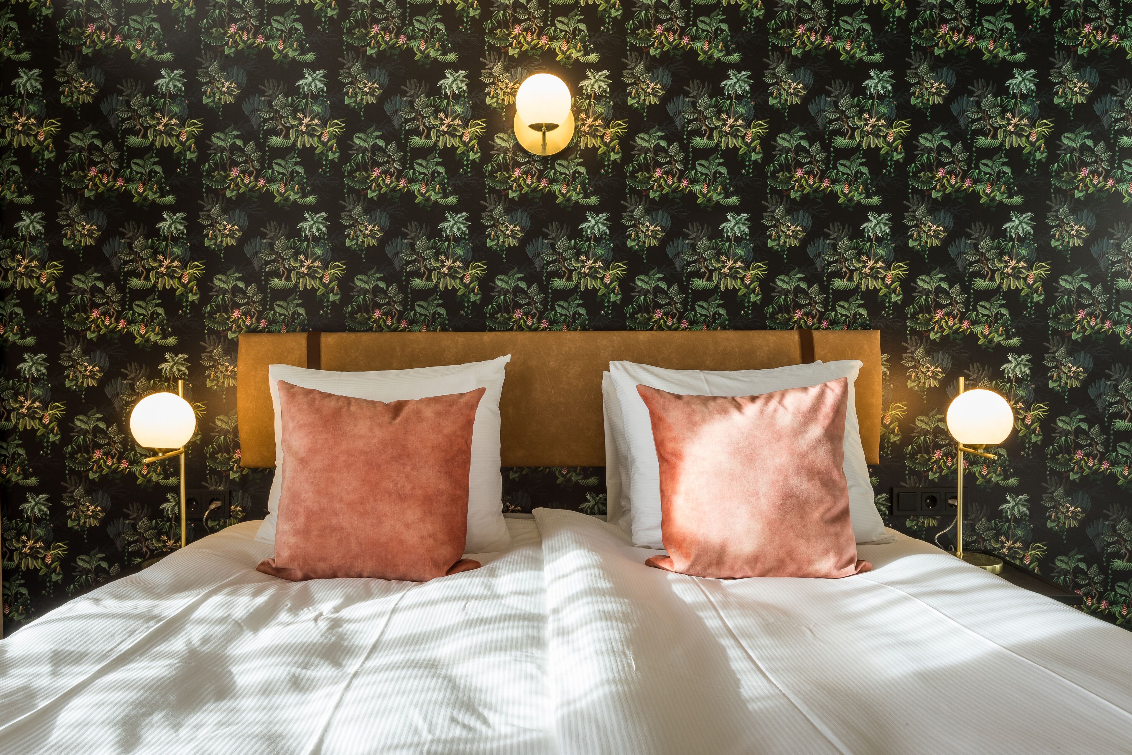 Cama com roupa de cama branca e duas almofadas cor-de-rosa, diante de uma parede com padrão de folhagem tropical.