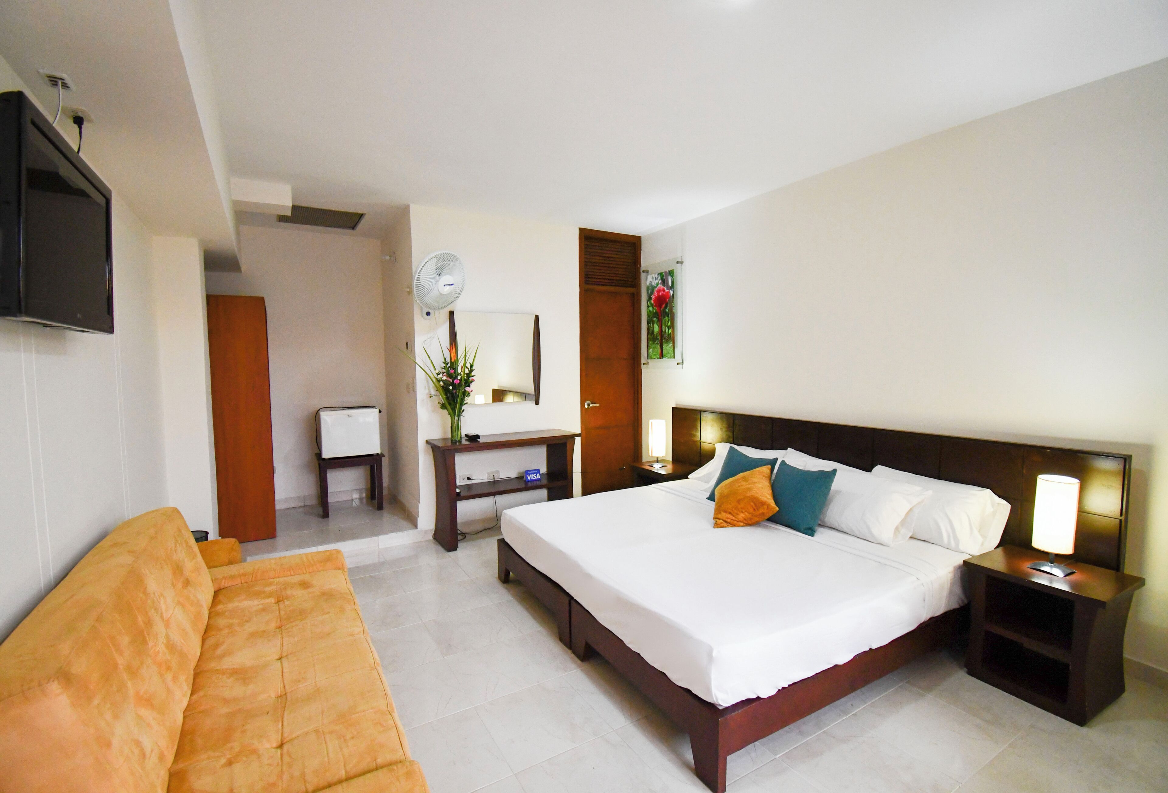 Quarto de hotel com uma cama grande, um sofá, uma escrivaninha com computador, uma televisão e um abajur.