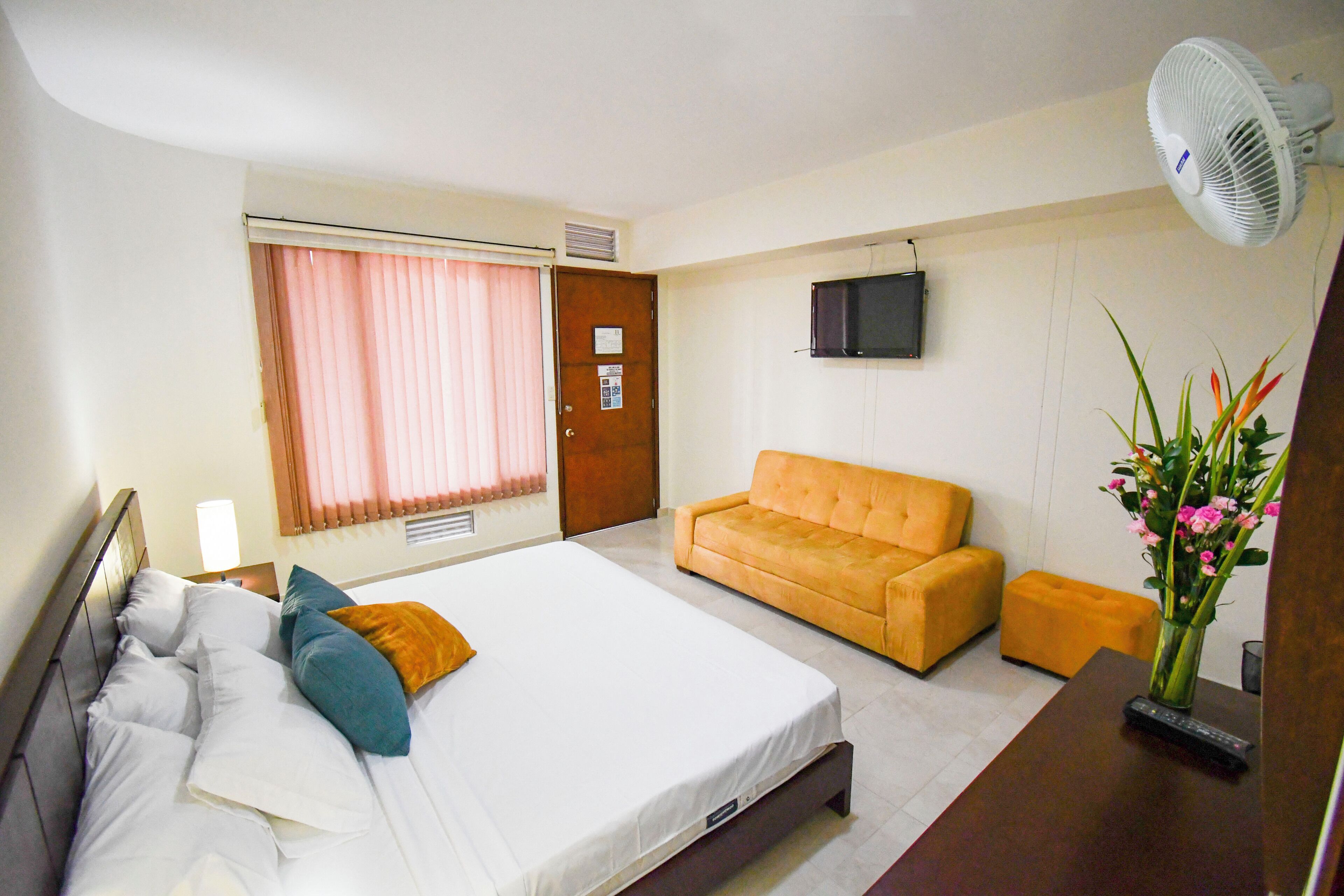 Quarto de hotel com cama, sofá, televisão, ventilador e um vaso com flores.