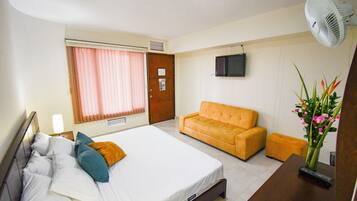 Quarto de hotel com cama, sofá, televisão, ventilador e um vaso com flores.