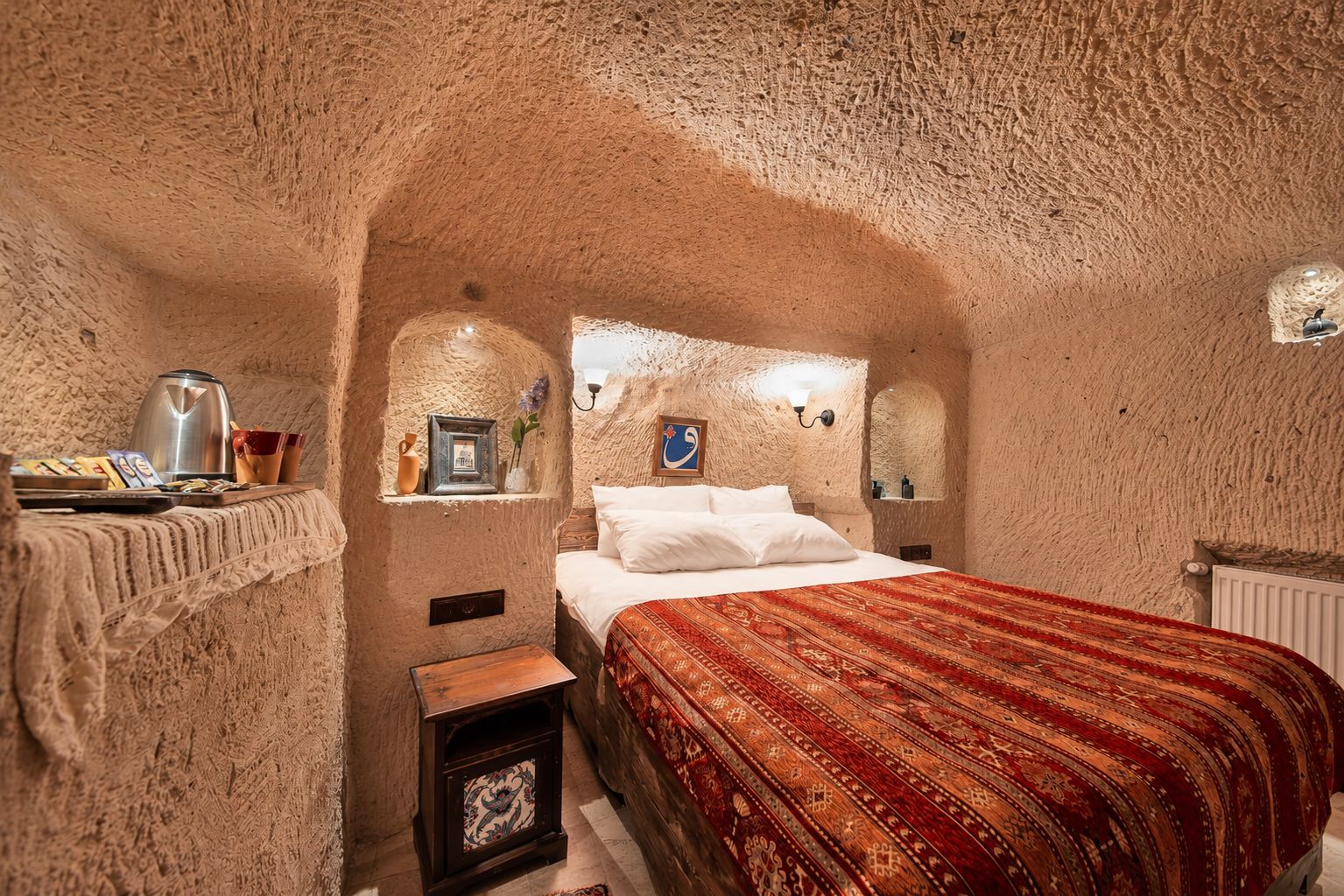 Um quarto aconchegante com uma cama, uma mesinha com chaleira e uma estante com objetos decorativos.
