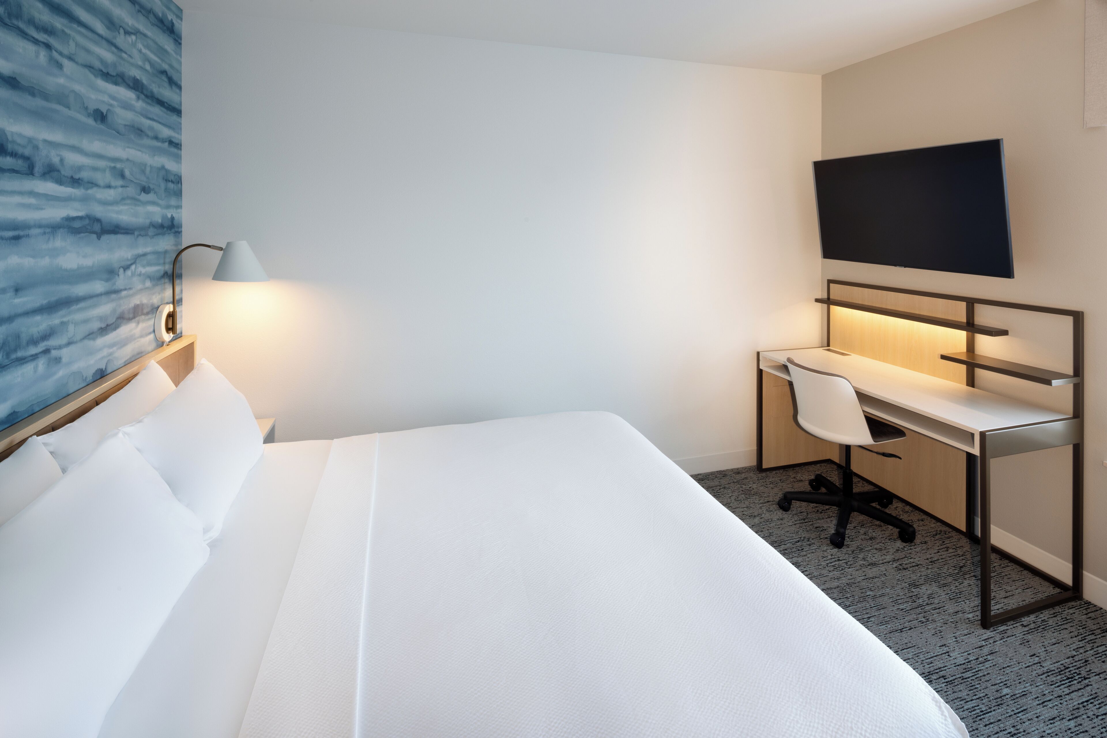 Quarto de hotel com cama, escrivaninha, cadeira, televisão e abajur.