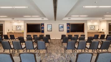 Sala de conferências com fileiras de cadeiras, um púlpito e duas telas grandes nas paredes.