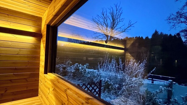 Uma cabana de madeira com janela que oferece vista para uma paisagem coberta de neve e uma árvore.