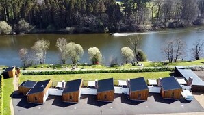Fileira de cabanas de madeira com telhados inclinados, dispostas linearmente ao longo de uma área pavimentada, próxima a um rio onde se vê um barco.