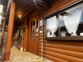 Uma cabana de madeira com varanda de pedra, uma janela grande com cortinas de renda e um vaso de planta no parapeito.