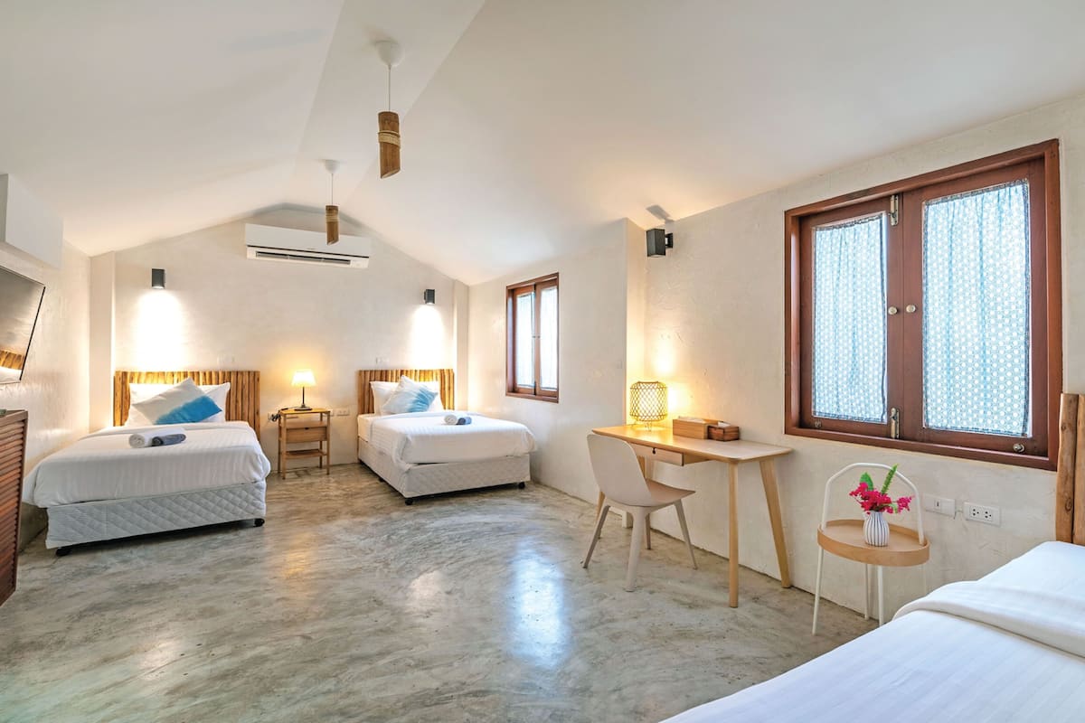 Quarto de hotel com duas camas, uma escrivaninha, uma cadeira e janela com cortinas estampadas.