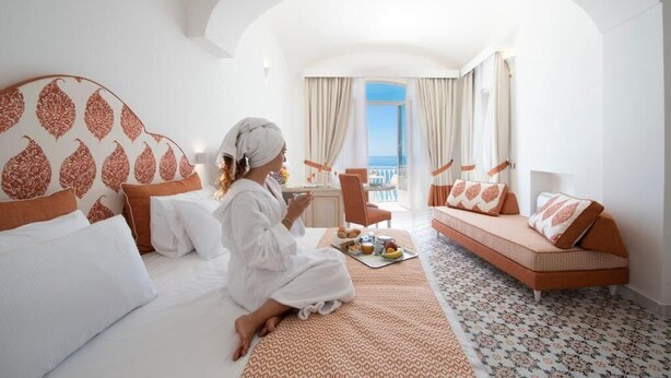 Quarto de hotel com uma cama grande, um sofá e uma área de refeições com vista para o mar.