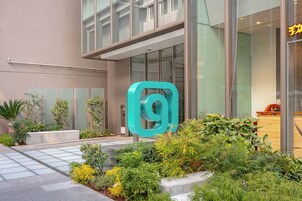 Um edifício moderno com um grande logotipo verde-turquesa, entrada de vidro e uma área de jardim bem cuidada.