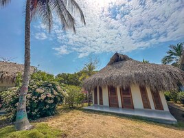 Um edifício com telhado de palha, varanda e venezianas de madeira, cercado por vegetação tropical e palmeiras.
