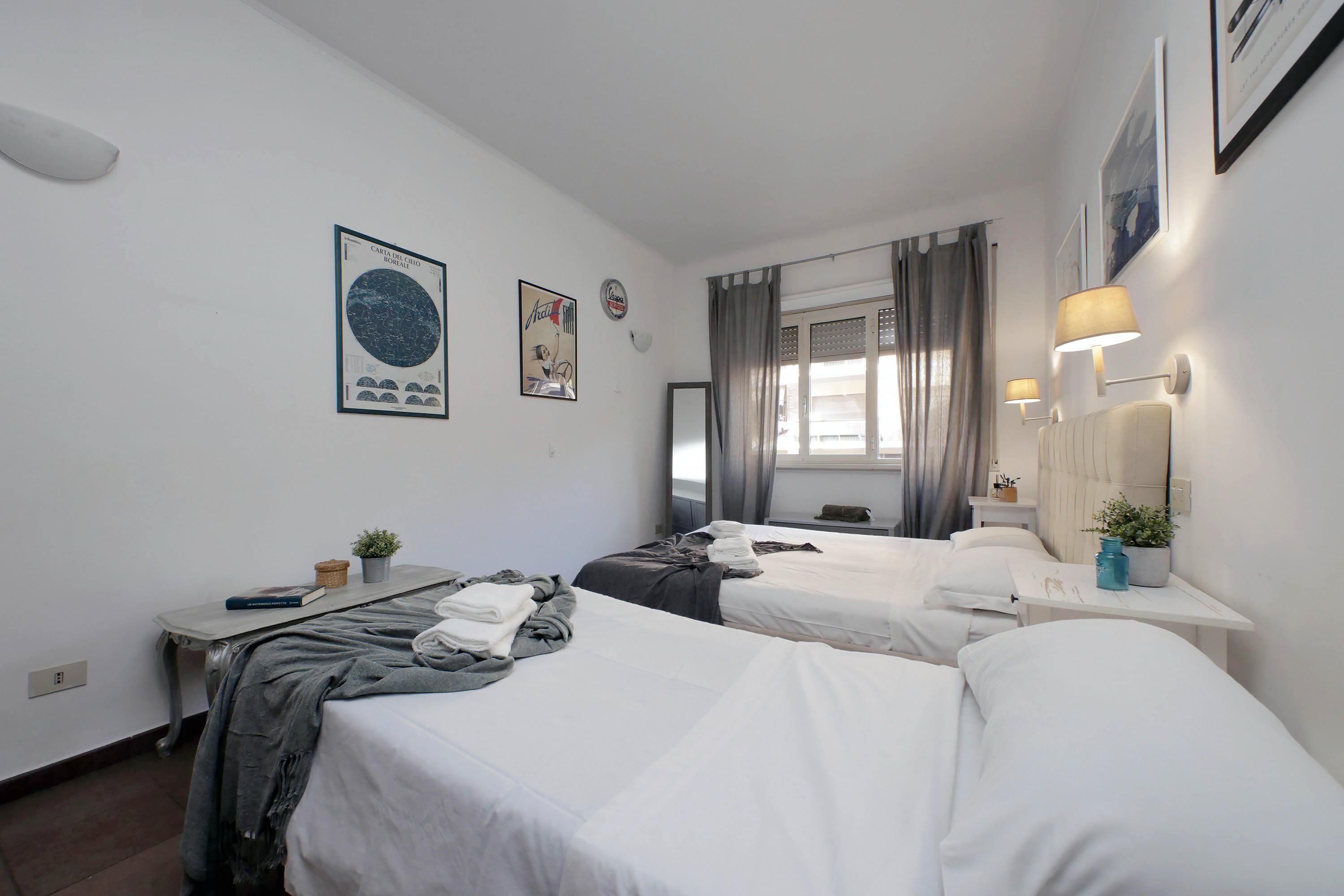 Quarto de hotel com duas camas, uma escrivaninha, janela com cortinas e luminárias na parede.