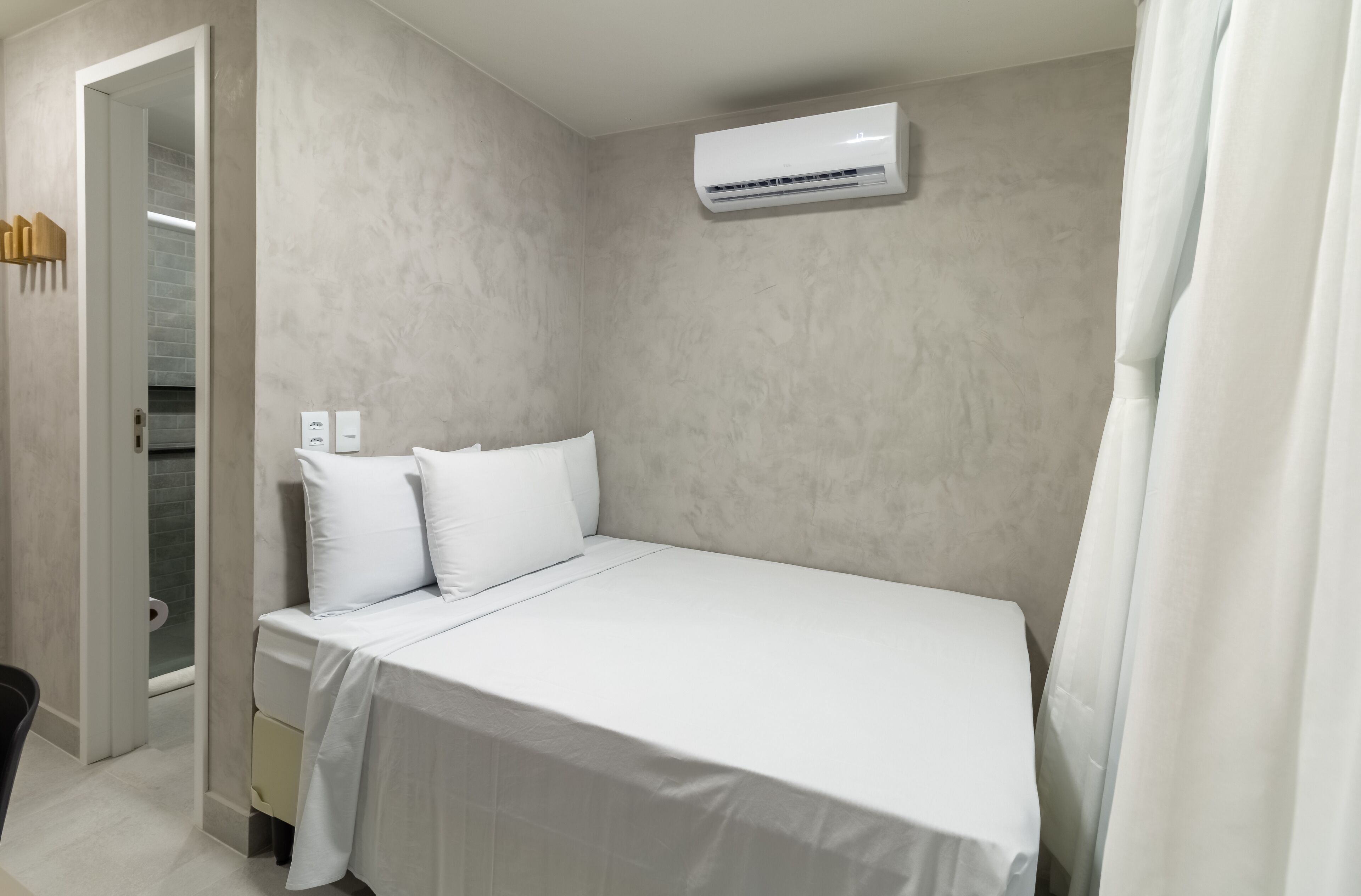 Um quarto pequeno e bem arrumado, com uma cama de solteiro, uma cortina branca e um ar condicionado na parede.