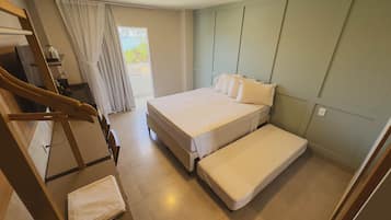 Quarto com cama, um banquinho, uma cadeira de madeira, uma mesinha e uma janela com cortinas.
