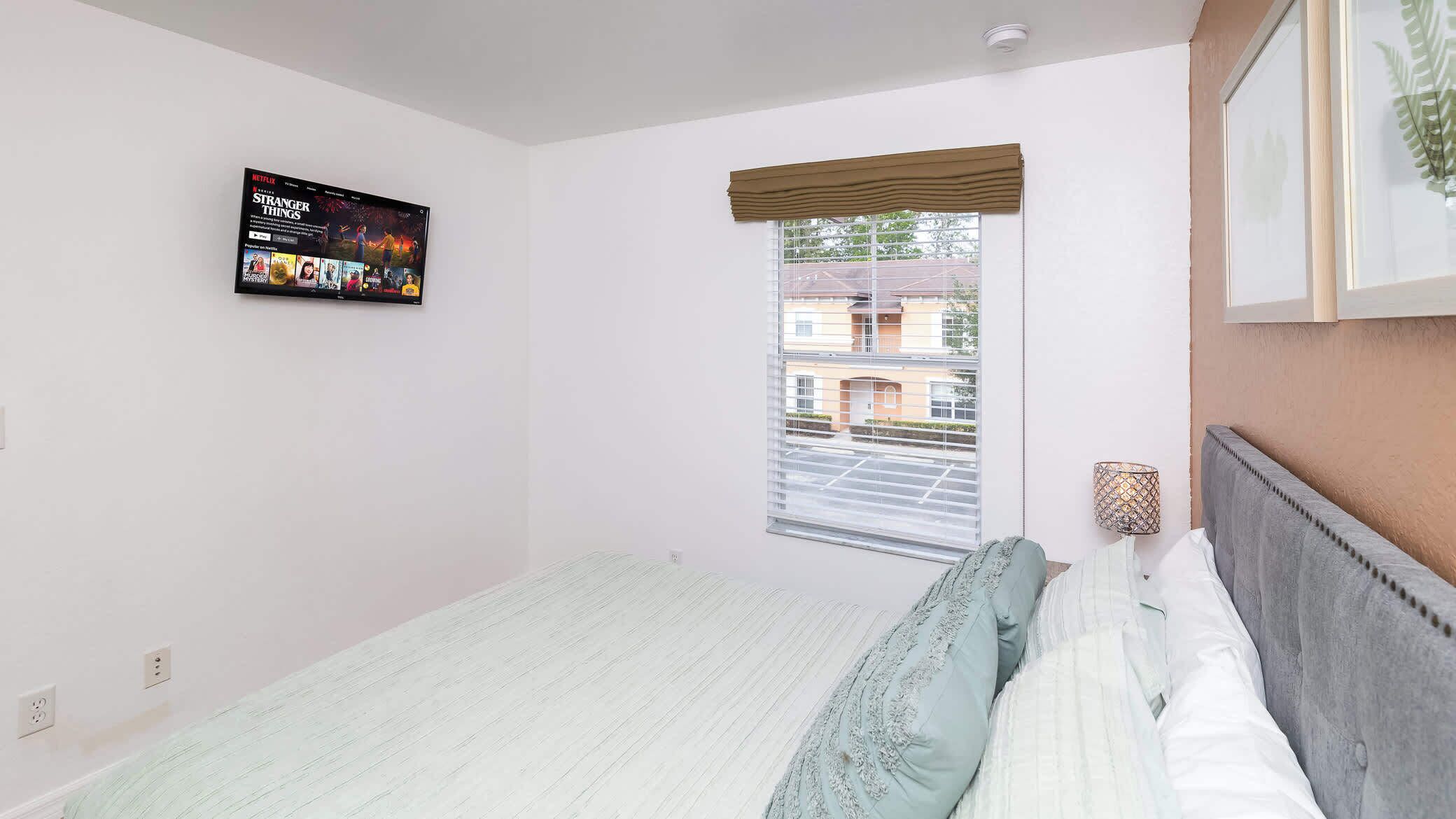 Quarto com cama, janela com persianas, TV fixada na parede exibindo um programa da Netflix e abajur na mesa de cabeceira.