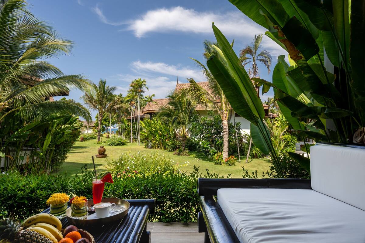 Vista de um jardim tropical a partir de uma varanda, com uma cama, um tabuleiro com frutas, uma bebida e um vaso de flores.