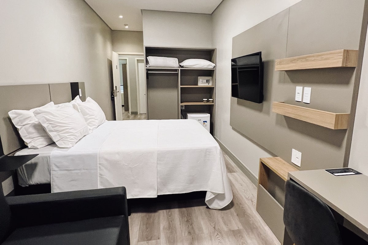 Quarto de hotel com uma cama, uma escrivaninha, uma cadeira, uma televisão e um guarda-roupa.