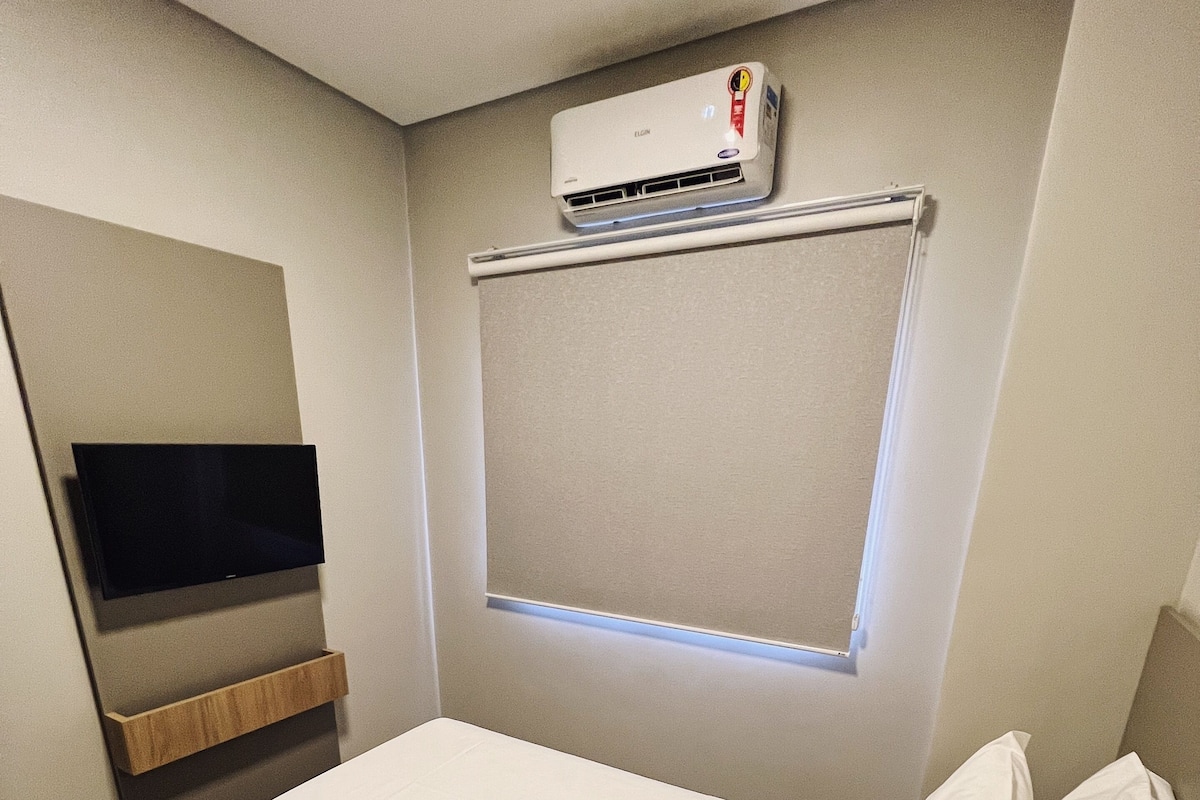 Quarto pequeno e bem arrumado, com uma cama, uma TV montada na parede e um aparelho de ar condicionado.