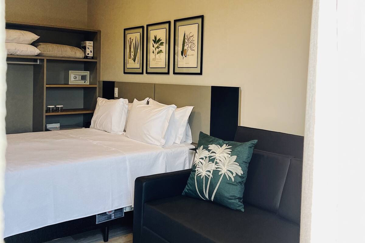 Quarto com cama, sofá preto, estante com almofadas e três impressões botânicas emolduradas na parede.
