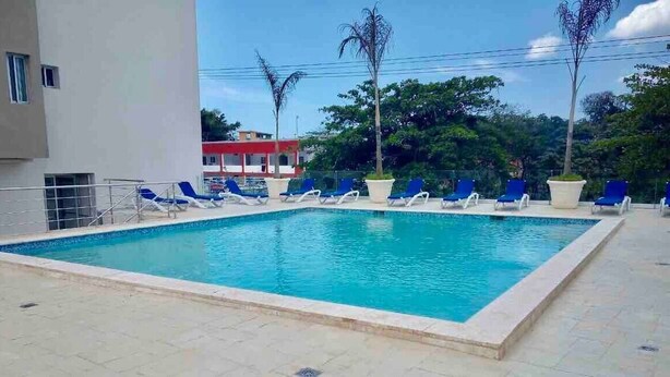 Uma piscina retangular com espreguiçadeiras azuis e plantas em vasos ao redor.