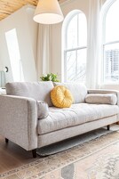 Um sofá cinza claro com uma almofada amarela, um tapete estampado e uma janela grande com cortinas.