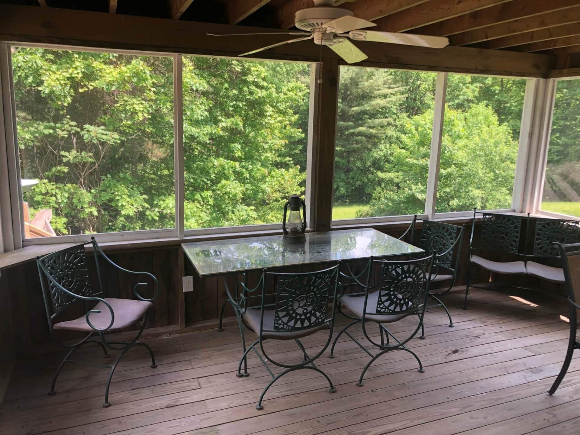 Varanda coberta com mesa de vidro e cadeiras de metal, grandes janelas com vista para uma área arborizada.