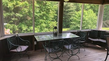 Varanda coberta com mesa de vidro e cadeiras de metal, grandes janelas com vista para uma área arborizada.