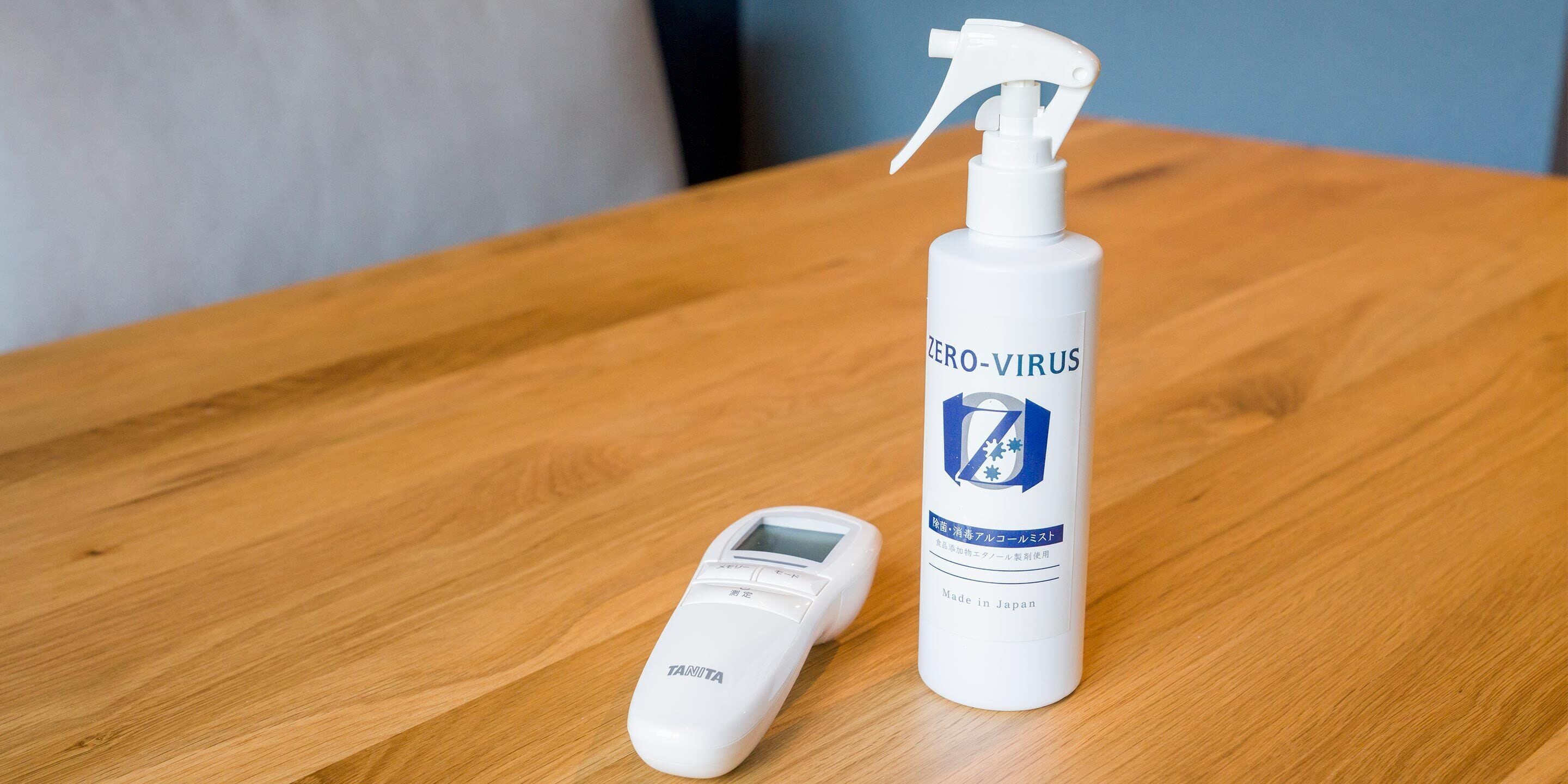 Um frasco spray branco com a etiqueta 'ZERO-VIRUS' e um dispositivo eletrônico branco sobre uma mesa de madeira.