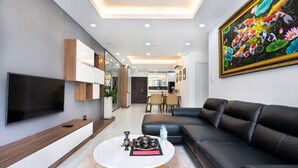 Uma sala de estar moderna com uma televisão de tela plana, um sofá de couro preto, uma mesa de centro branca e uma área de jantar ao fundo.