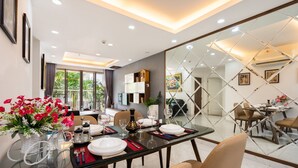 Uma sala de jantar com mesa de vidro posta para quatro pessoas, rodeada por cadeiras. Há um vaso com flores sobre a mesa, e o ambiente tem design moderno com um espelho grande e vista para o exterior.