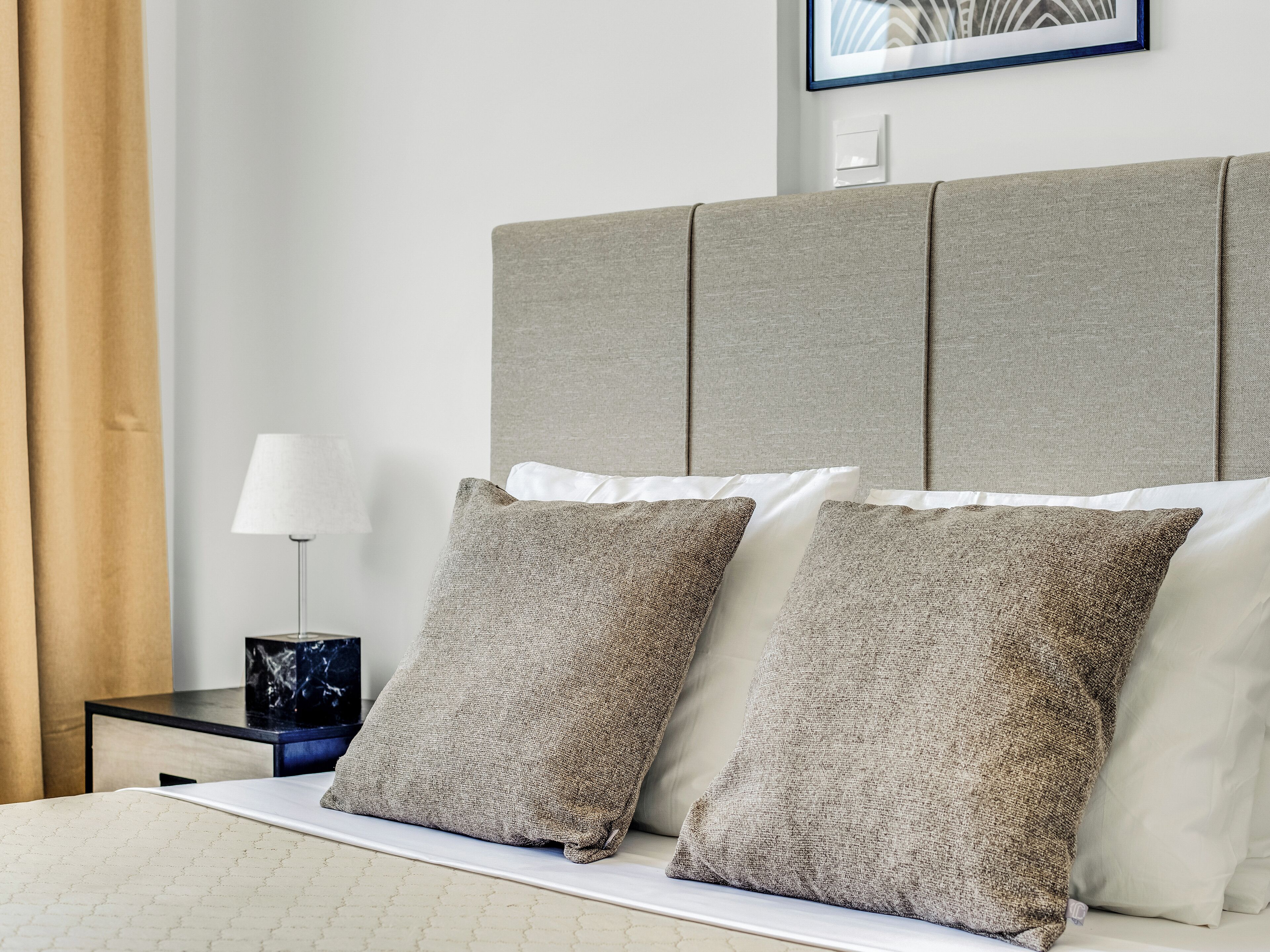 Uma cama bem arrumada com cabeceira, uma mesa de cabeceira com abajur e um quadro na parede.