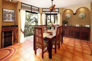 Uma sala de jantar com mesa e cadeiras de madeira, um ventilador de teto, vista para palmeiras e uma cozinha com armários de madeira.