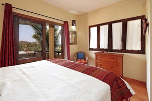 Um quarto com cama, cômoda de madeira, cadeira, janela com cortinas e vista para palmeiras.