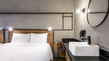 Quarto de hotel com cama, escrivaninha com pia, uma cadeira e espelho.