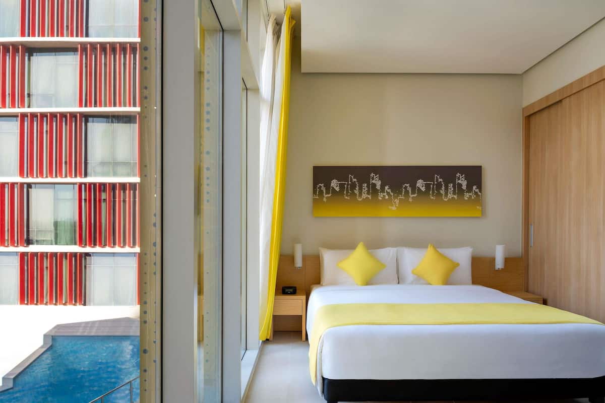 Quarto de hotel moderno com uma cama grande, uma parede de destaque amarela e uma pintura de paisagem urbana.